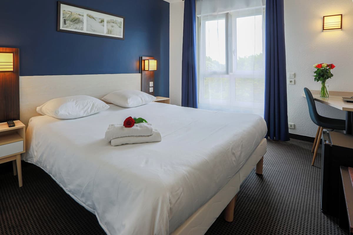 Quarto de hotel com uma cama grande, uma escrivaninha e uma cadeira. Há uma janela com cortinas, uma luminária na parede e um quadro emoldurado acima da cama.