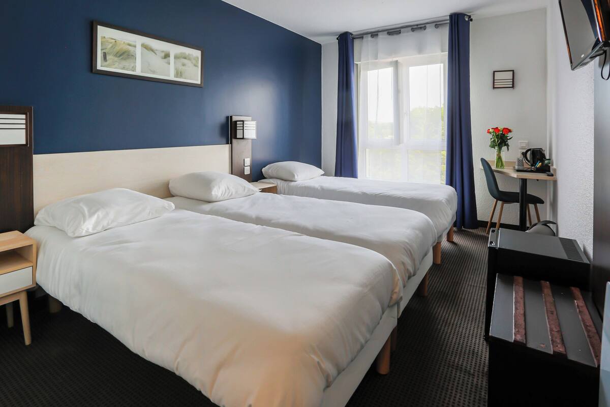 Quarto de hotel com duas camas, uma secretária com cadeira, televisão e janela com cortinas.