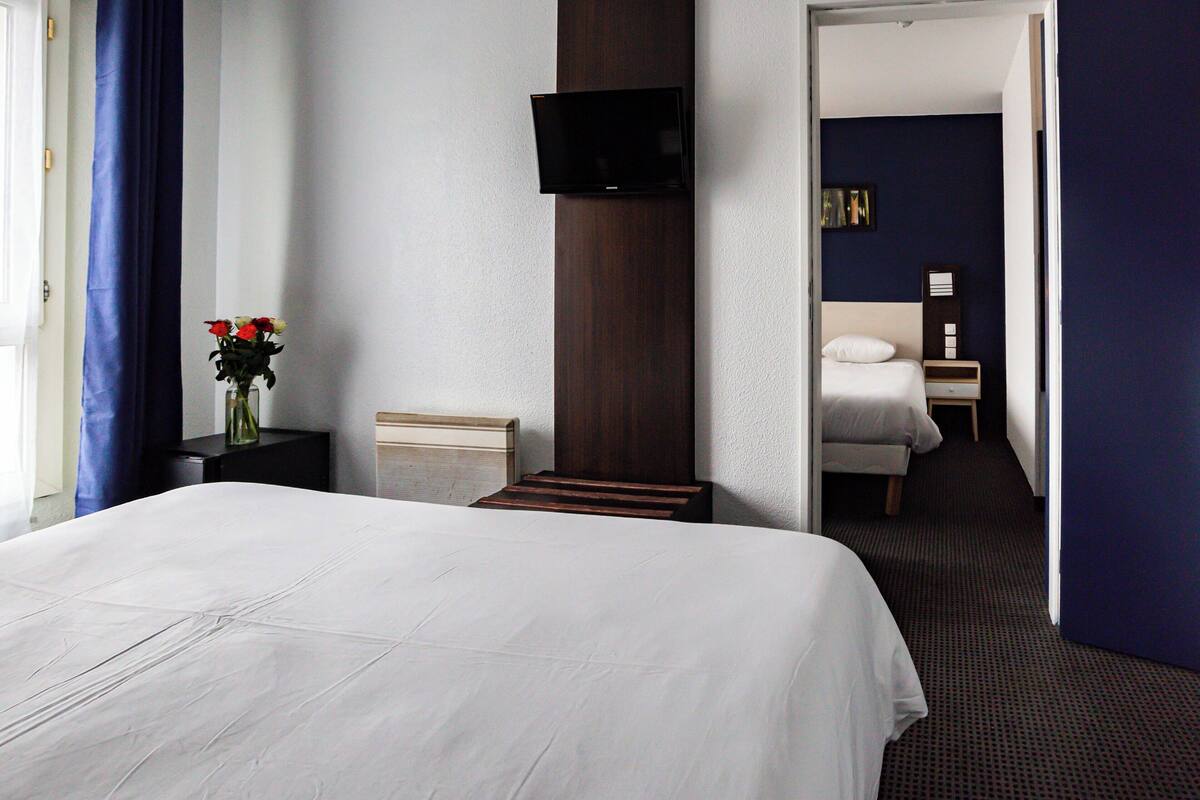 Quarto de hotel com uma cama grande, televisão de tela plana na parede, mesa de cabeceira com abajur e uma pequena mesa com um vaso de flores.