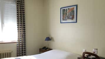 Quarto de hotel com cama, escrivaninha com cadeira, abajur, quadro na parede e janela com cortinas.