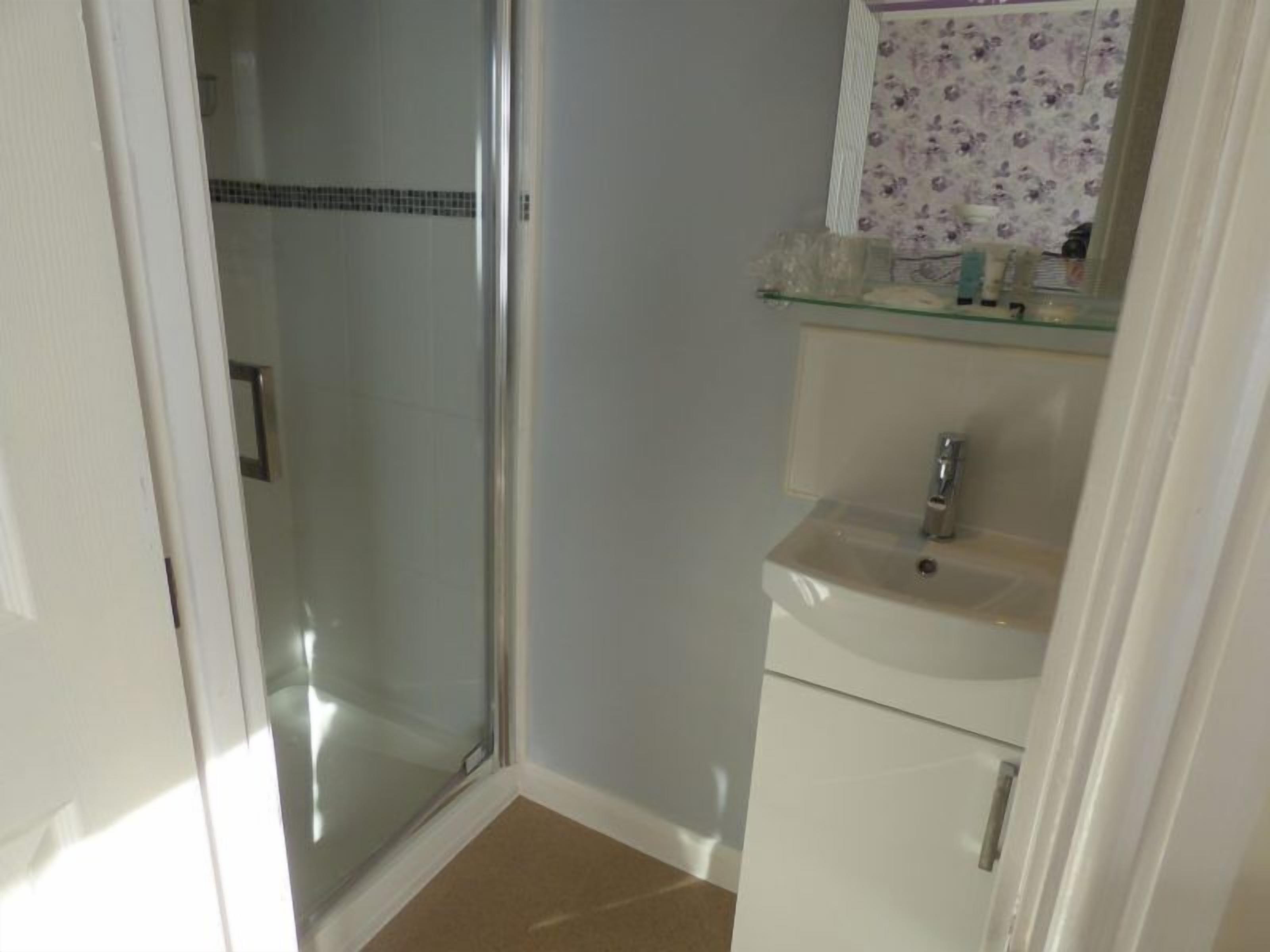 Uma casa de banho com cabine de duche em vidro, lavatório com espelho e uma pequena estante com artigos de higiene pessoal.