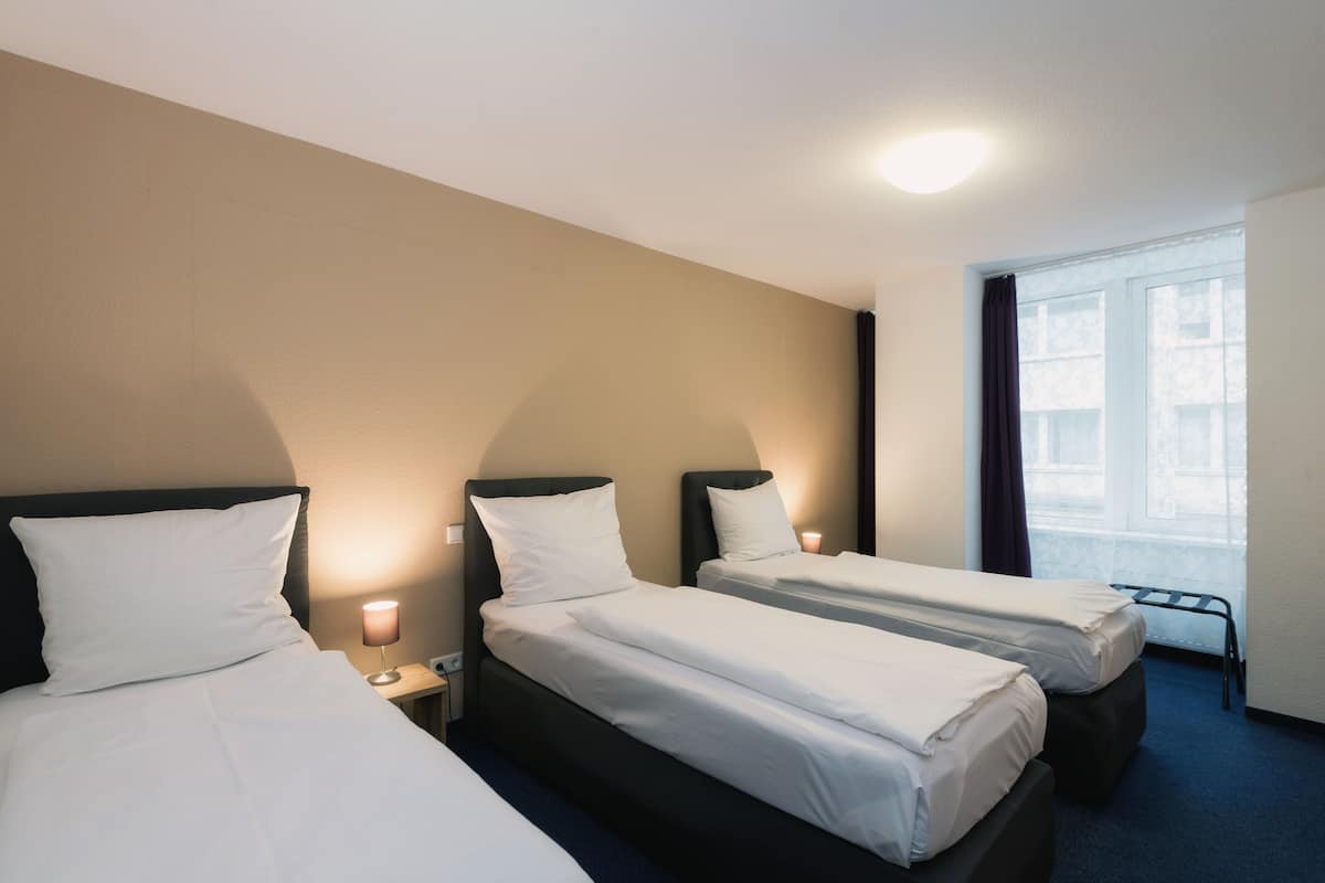 Quarto de hotel com duas camas de solteiro, uma mesa de cabeceira com abajur, uma janela com vista e uma luminária no teto.