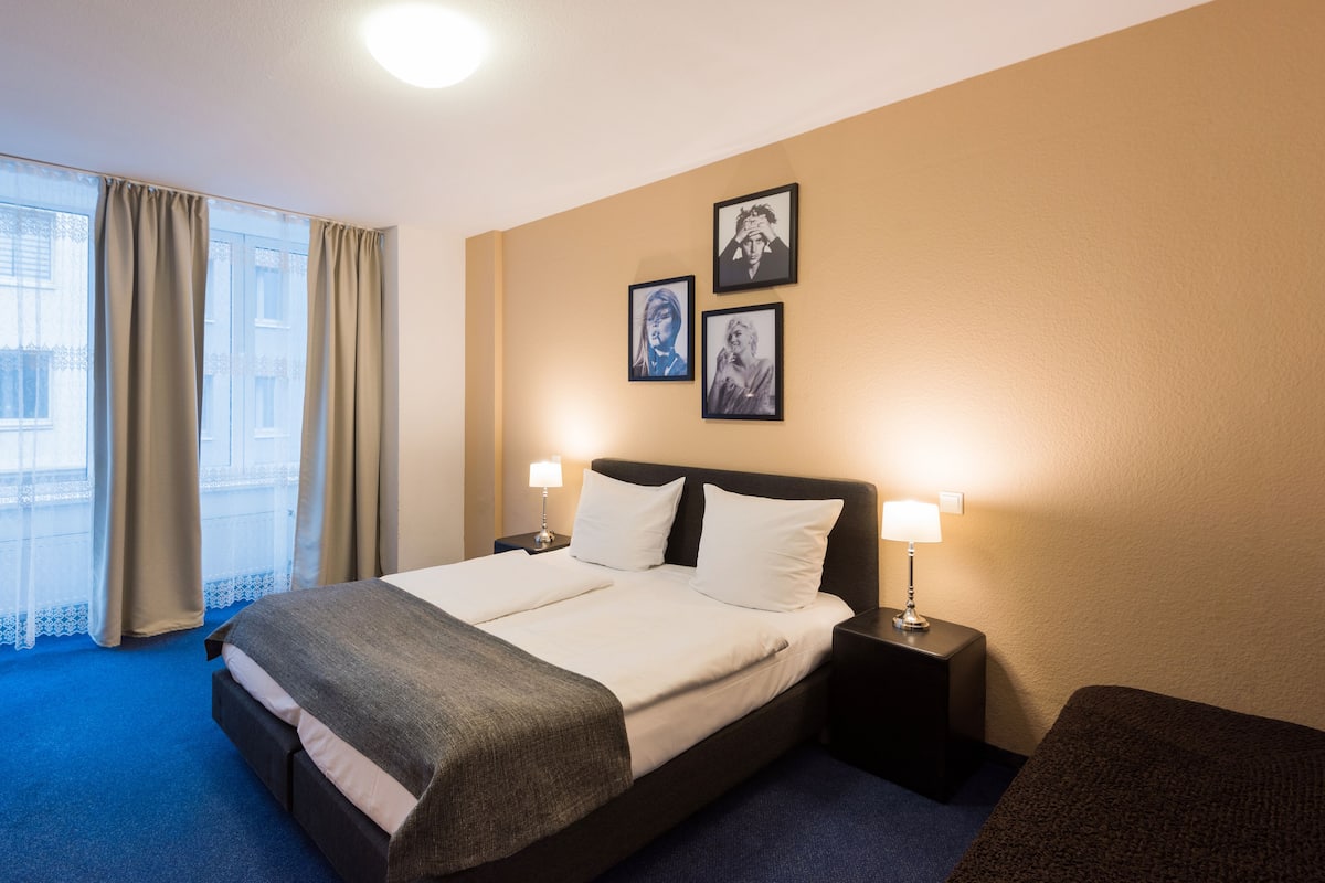Quarto de hotel com uma cama grande, duas mesas de cabeceira com luminárias, uma janela com cortinas e três quadros na parede.
