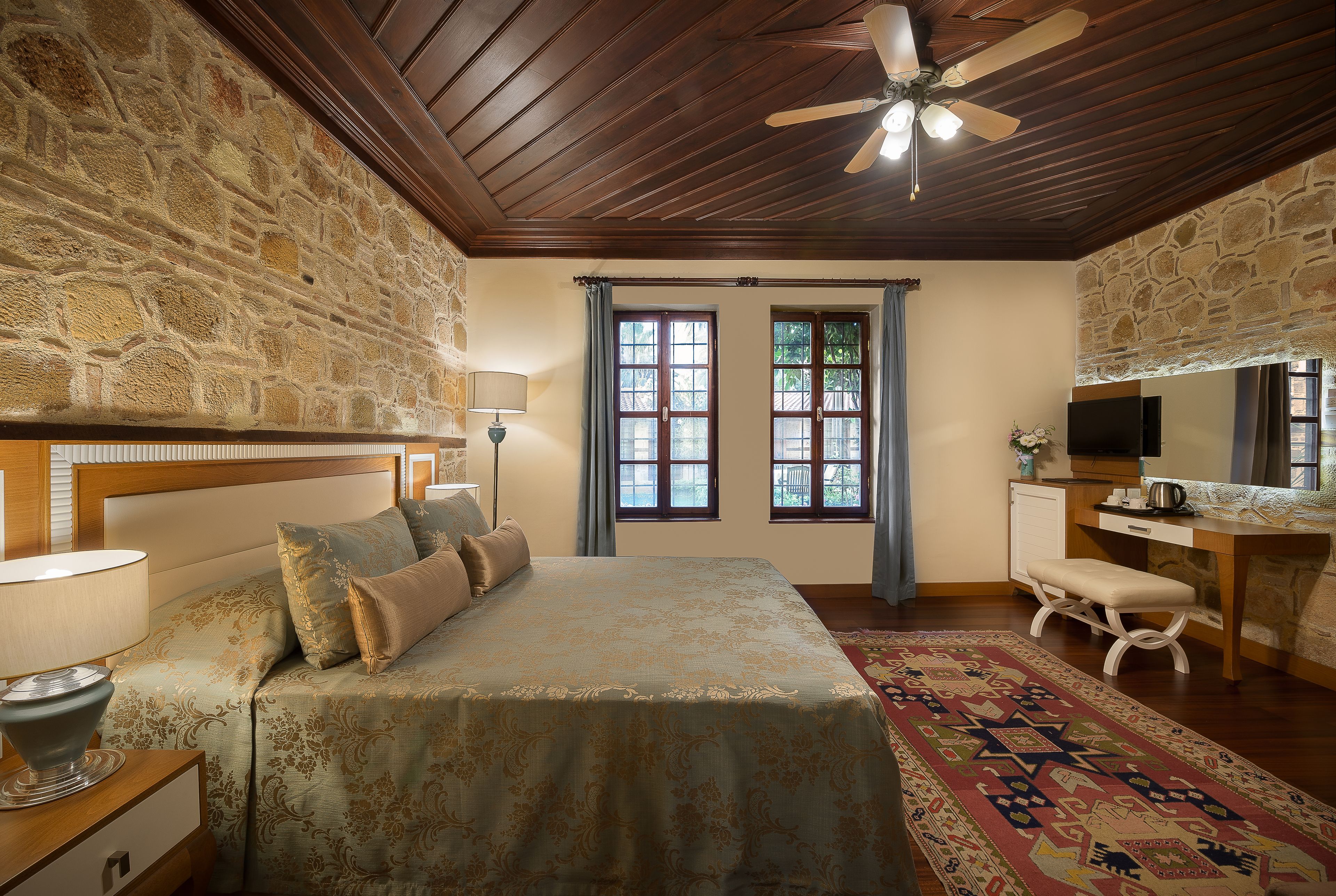 Quarto com parede de pedra, teto de madeira, cama com colcha estampada, escrivaninha com televisão e janela com cortinas azuis.