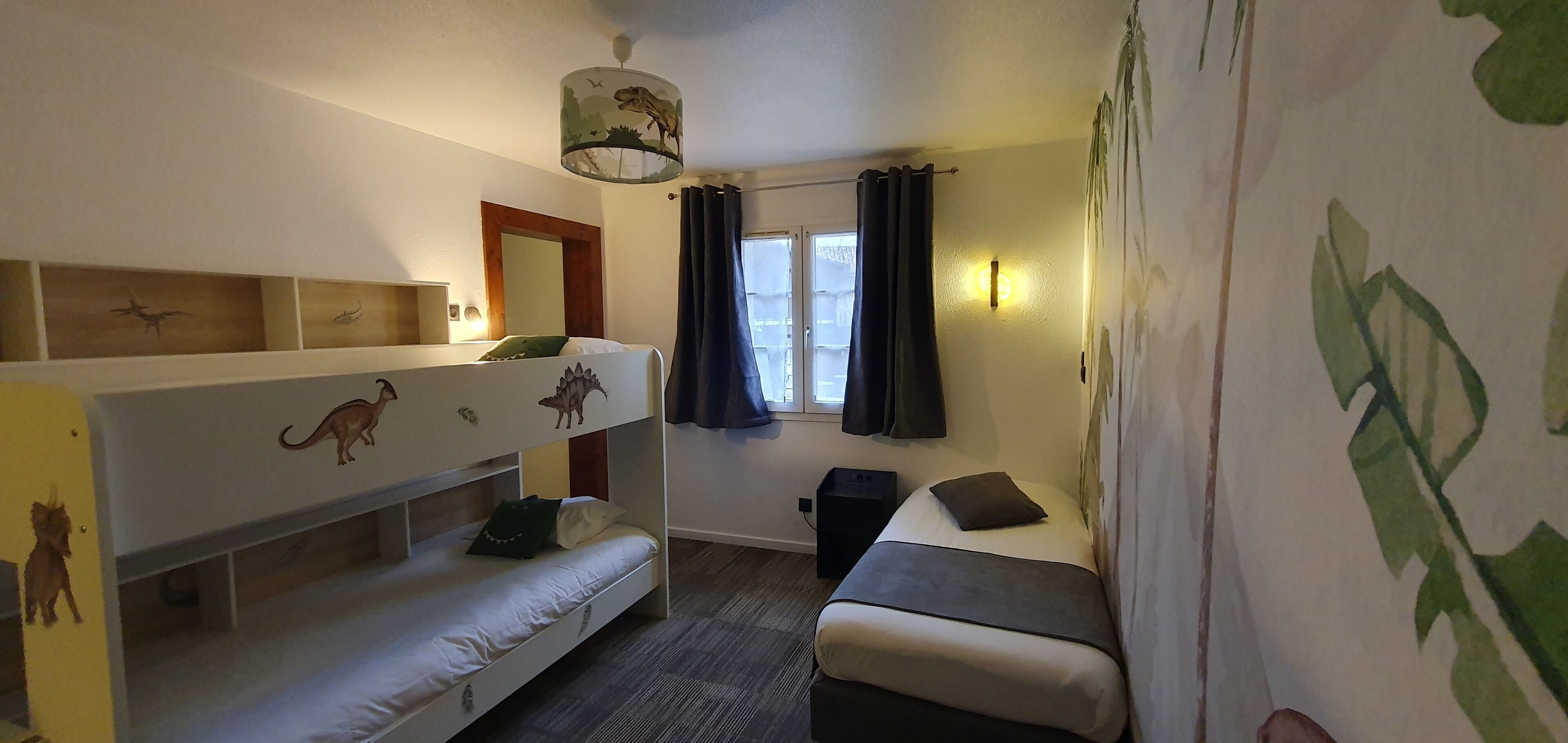Quarto em dormitório com beliches, uma escrivaninha, uma cadeira, janela com cortinas e luminária fixa na parede.