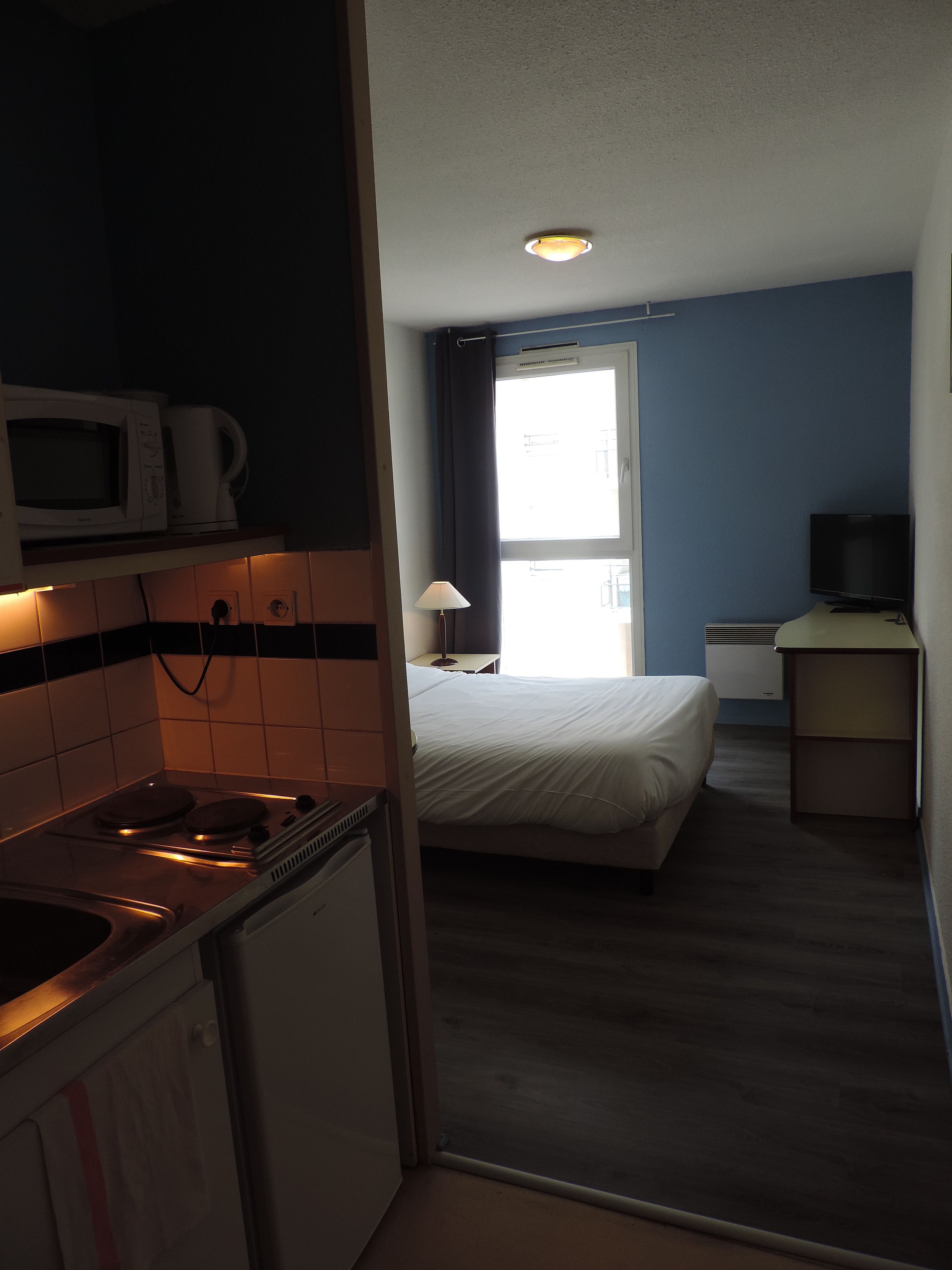 Quarto de hotel compacto com cozinha compacta, uma cama, escrivaninha e televisão.