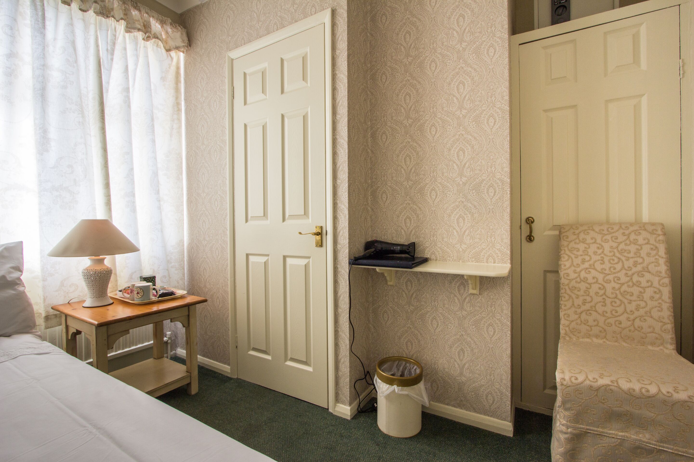 Quarto de hotel com cama, mesa de cabeceira com abajur, cadeira, porta, lixeira e prateleira com um livro.