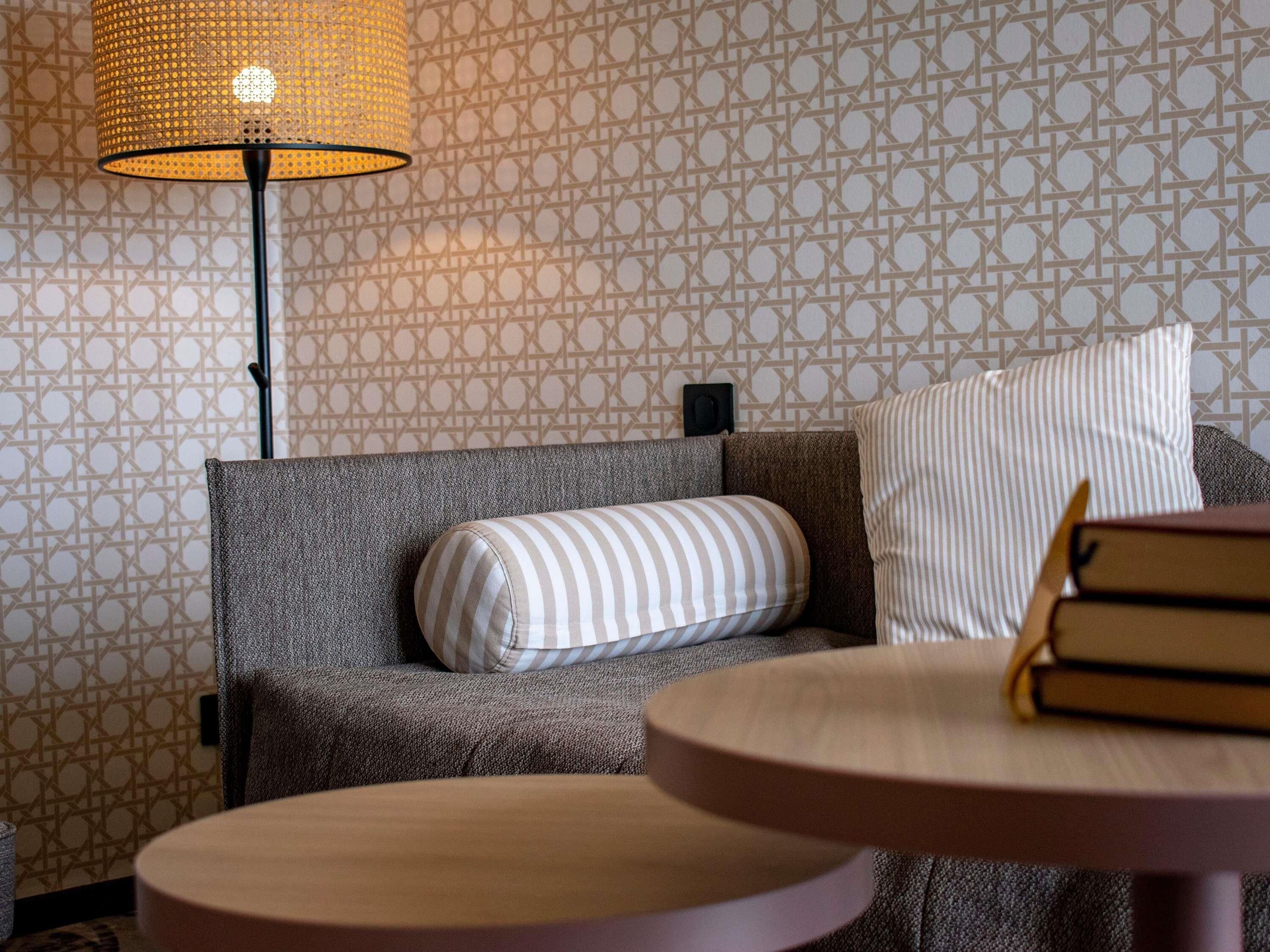 Quarto com papel de parede estampado, sofá com almofadas listradas, luminária de chão e mesa redonda com livros.