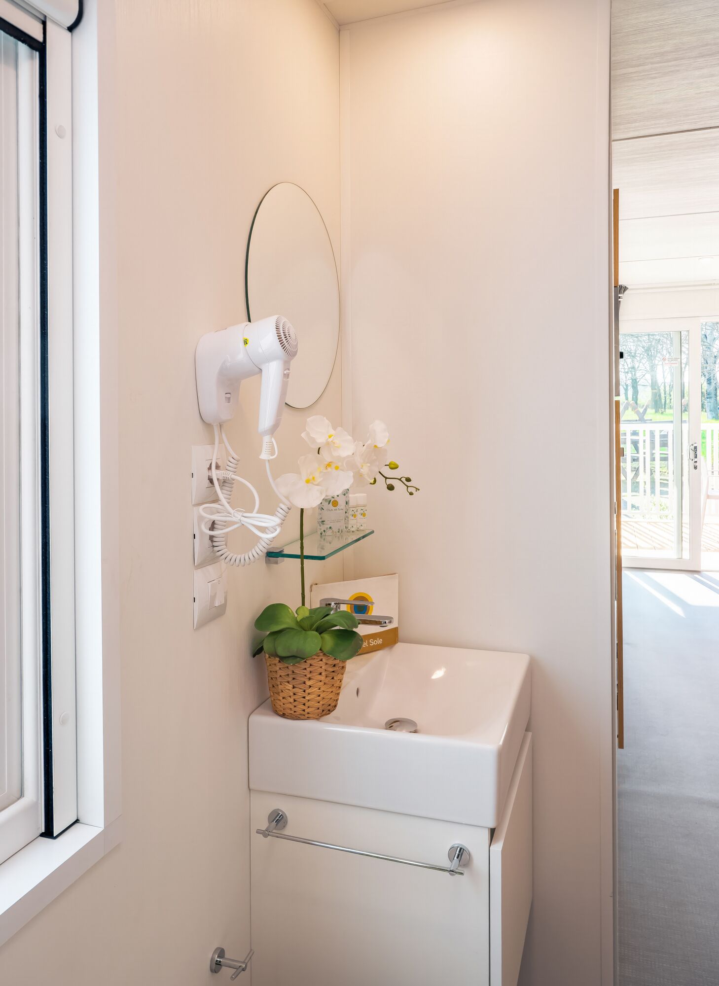 Uma casa de banho com uma pia branca, um secador de cabelo fixado na parede, uma pequena prateleira com artigos de higiene pessoal e uma planta em vaso.