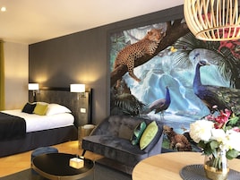 Quarto de hotel com cama, sofá, mesa de centro e um grande mural na parede com um leopardo e um pavão.