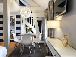 Quarto de hotel moderno com uma mesa de jantar redonda, cadeiras brancas, um vaso com flores brancas, um abajur e uma abacaxi decorativa sobre uma cômoda branca.