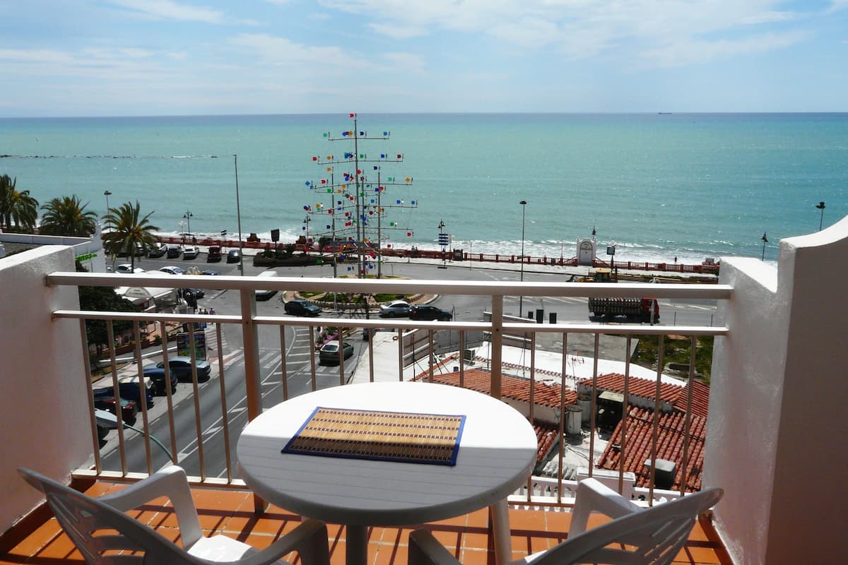 Varanda com mesa e cadeiras, vista para a orla marítima, mar e uma construção colorida ao longe.