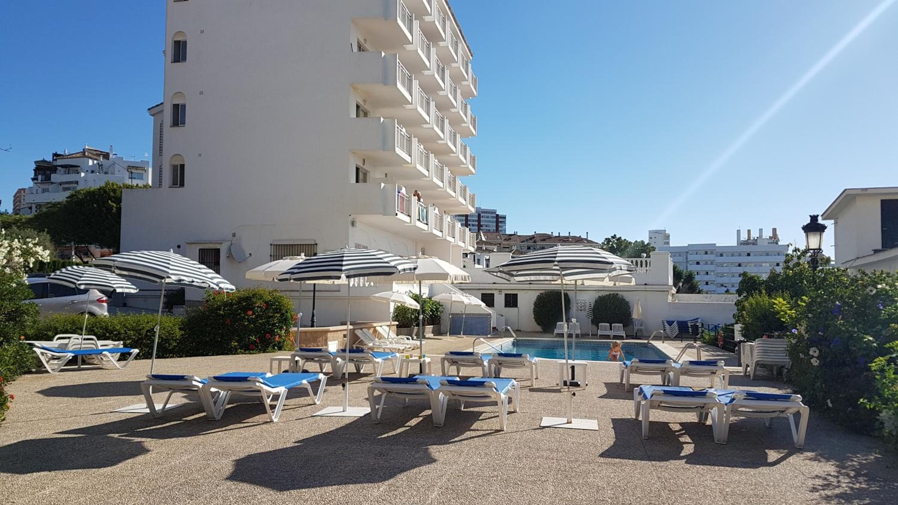 Uma área ao lado da piscina com espreguiçadeiras e guarda-sóis, piscina e um edifício branco com varandas.