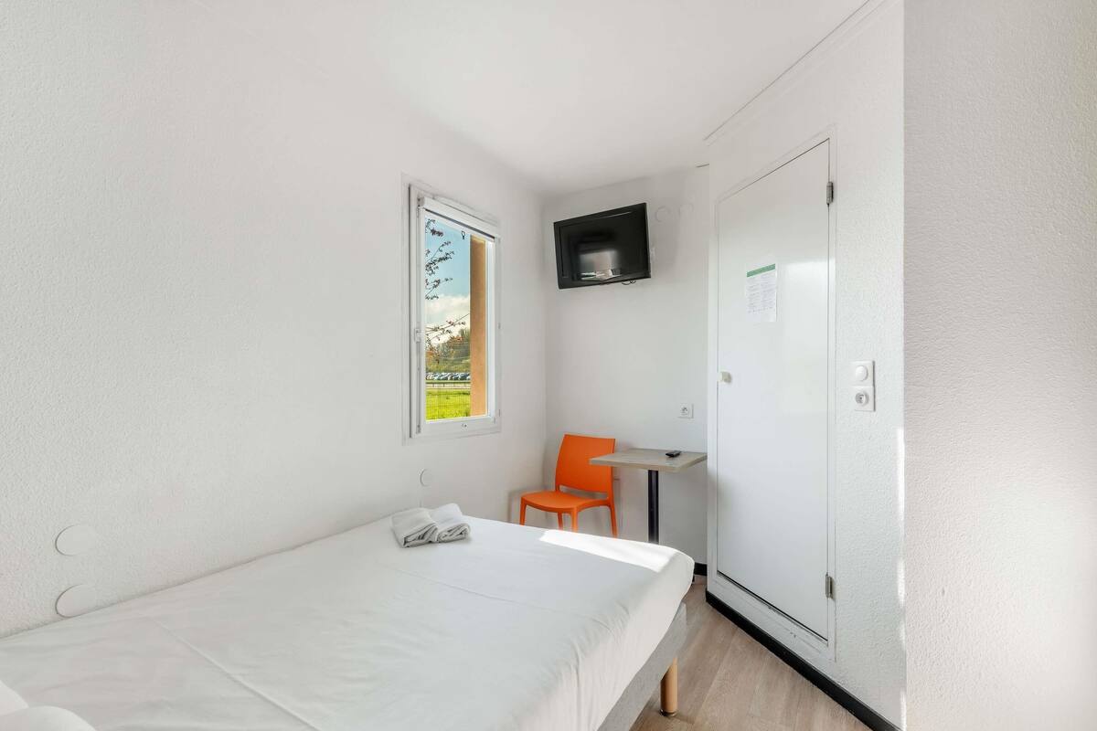Quarto pequeno com cama de solteiro, janela, uma cadeira, uma mesinha e televisão fixada na parede.