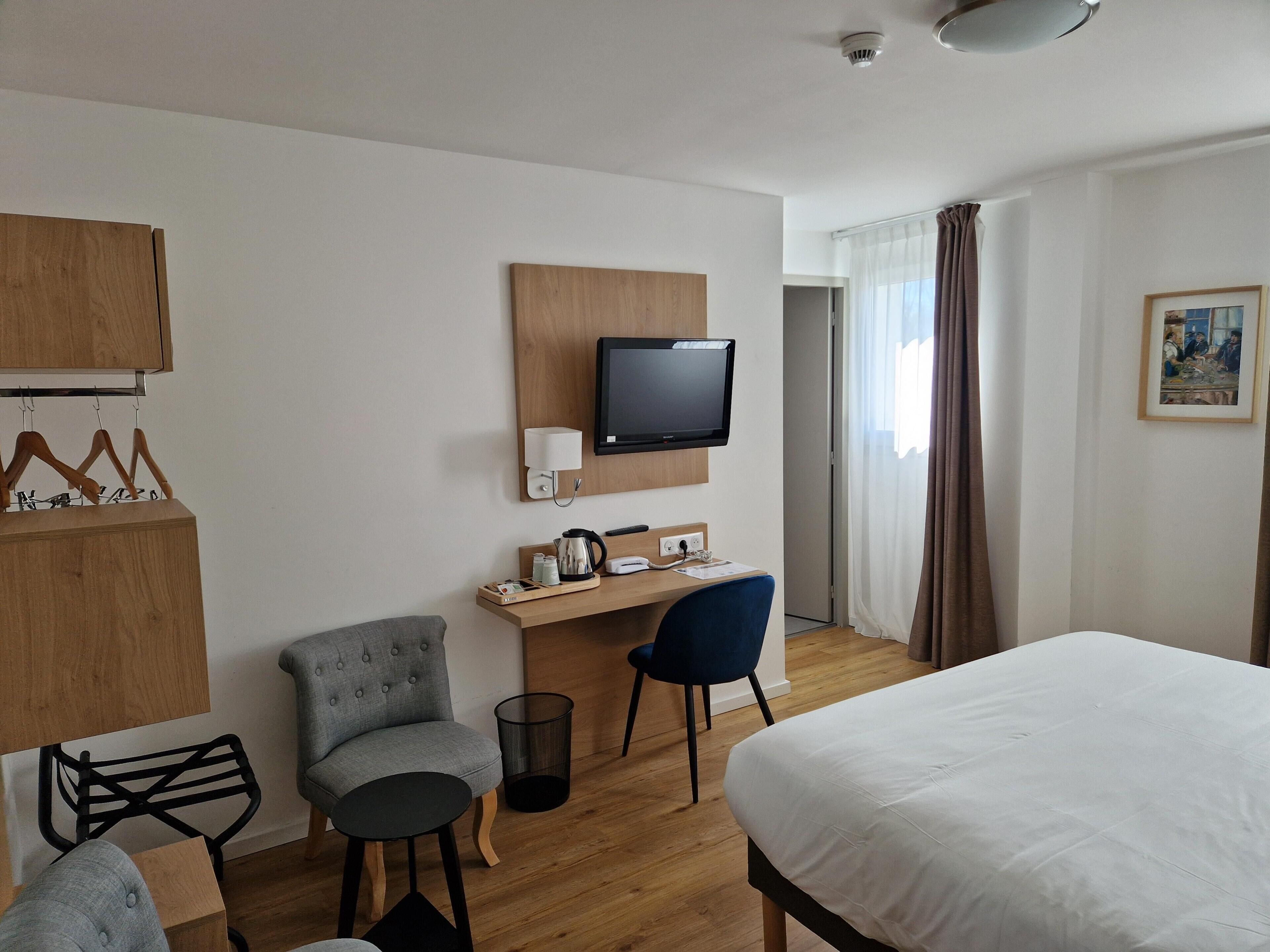 Quarto de hotel com cama, escrivaninha com cadeira, televisão e guarda-roupa.