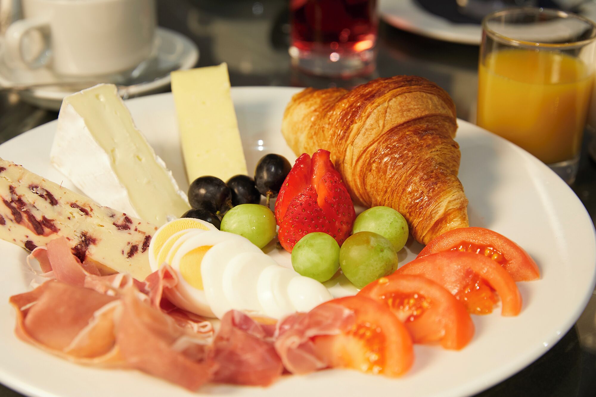 Prato de café da manhã com queijo, presunto, ovos, uvas, morangos, tomates e um croissant.