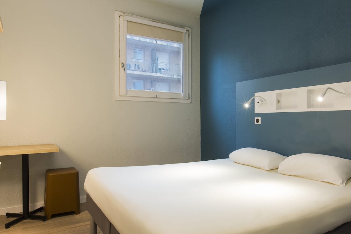 Quarto de hotel com cama, escrivaninha, um banquinho pequeno, janela e luminária na parede.