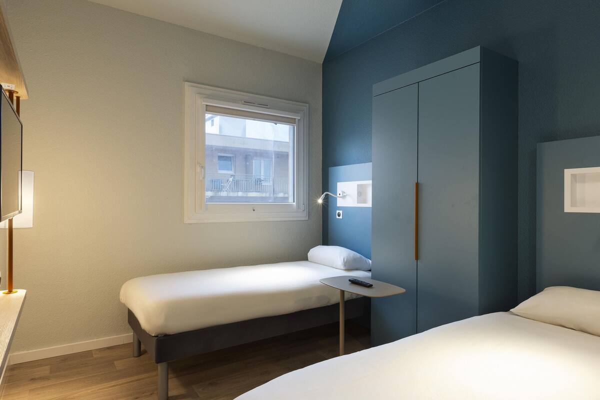Quarto de hotel com uma cama, uma mesinha, um guarda-roupa, uma janela e um abajur.