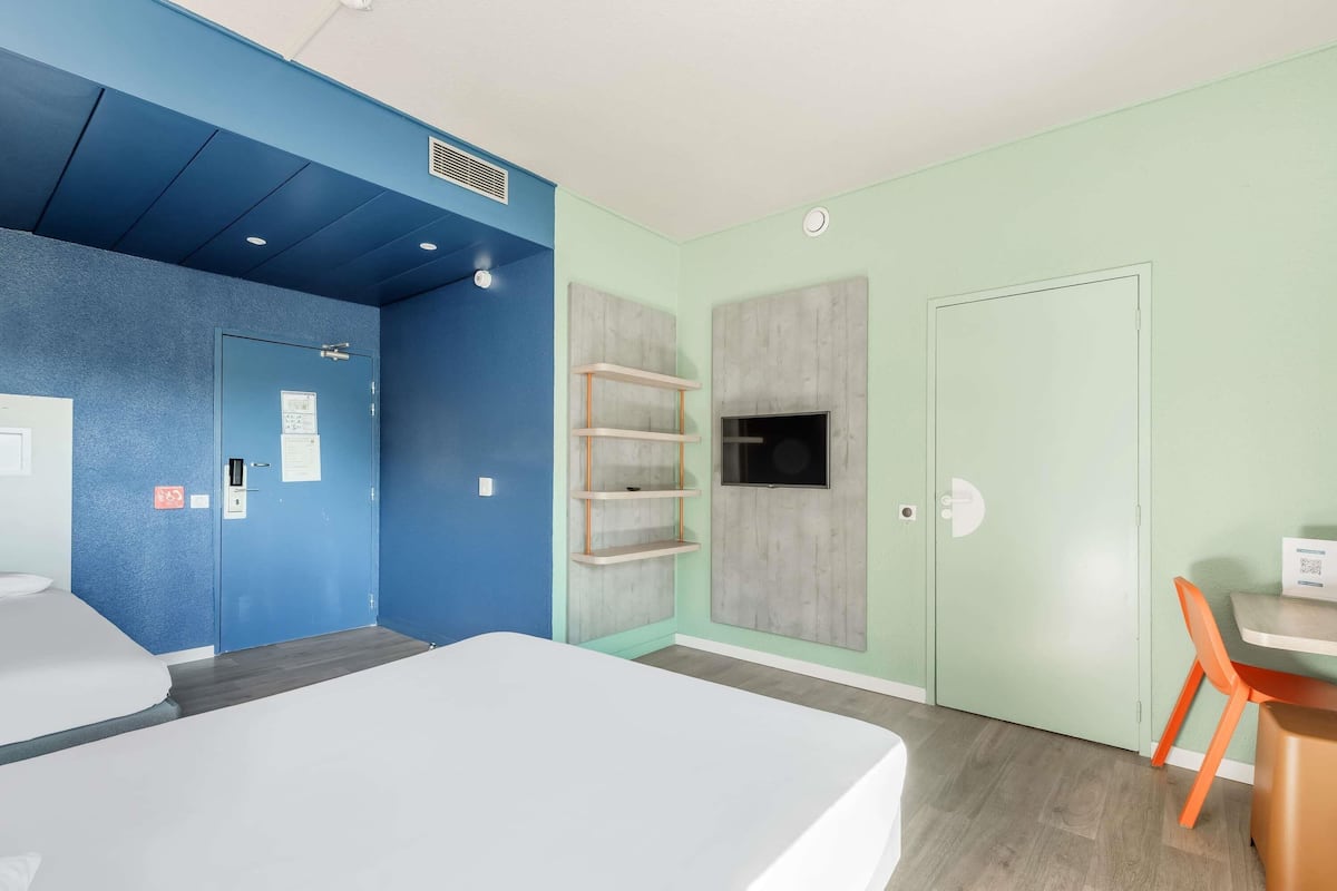 Quarto de hotel com cama, escrivaninha, televisão fixada na parede e porta com puxador.
