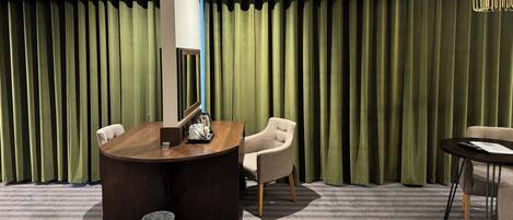 Quarto de hotel com uma escrivaninha de madeira, duas cadeiras, um cesto de lixo e uma janela grande com cortinas verdes.