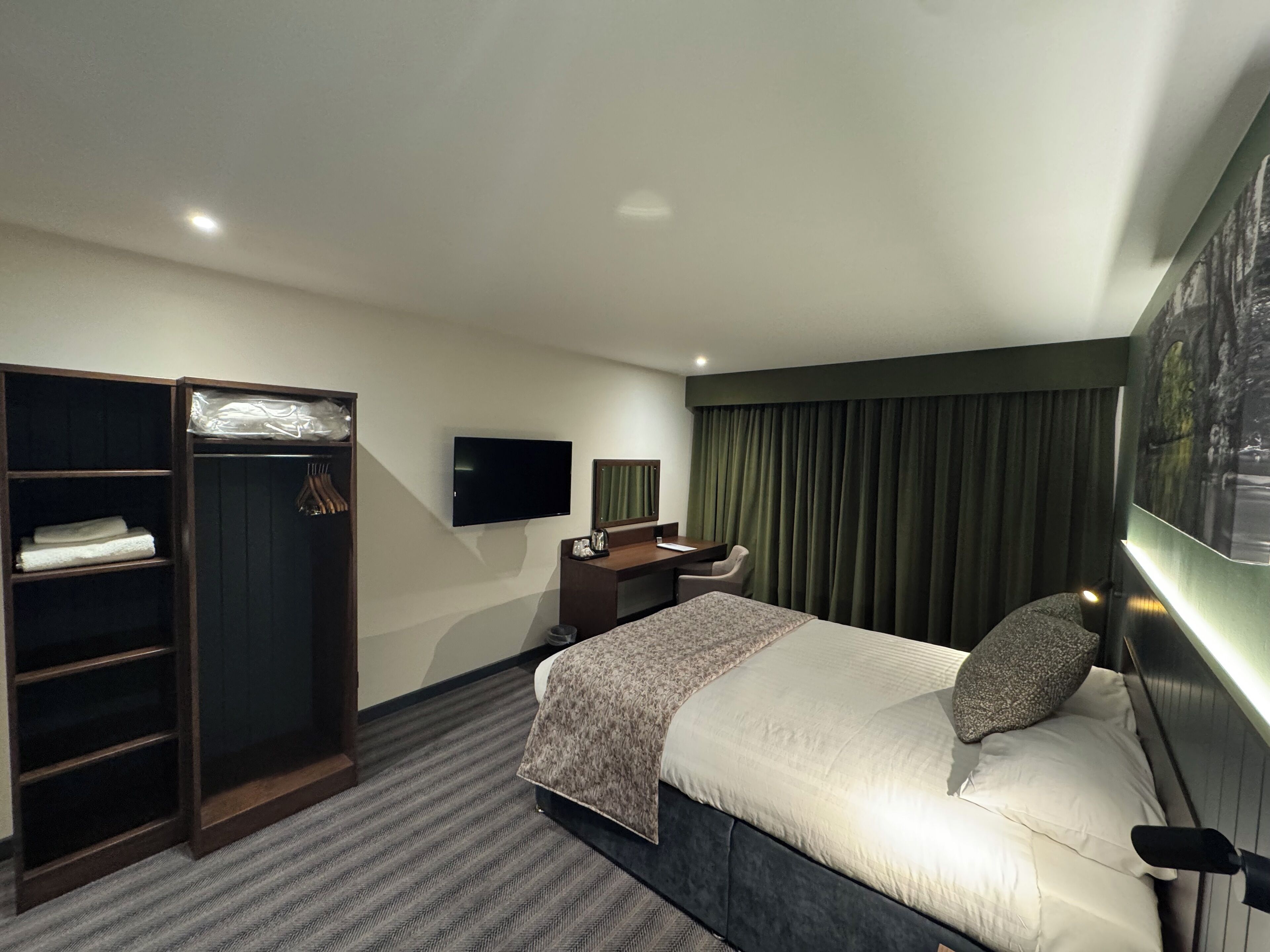 Quarto de hotel com cama, escrivaninha, televisão, guarda-roupa e uma janela grande com cortinas.