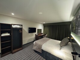 Quarto de hotel com cama, escrivaninha, televisão, guarda-roupa e uma janela grande com cortinas.