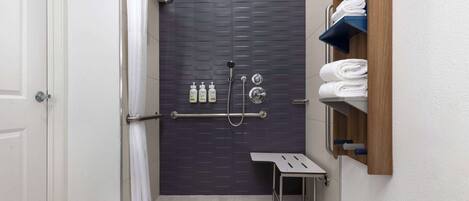 Um espaço de chuveiro estreito com parede revestida de azulejos escuros, uma porta branca, um porta-toalhas com toalhas dobradas e uma pequena mesa.