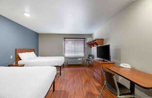 Quarto de hotel com duas camas, uma mesa, uma televisão e uma janela com persianas.