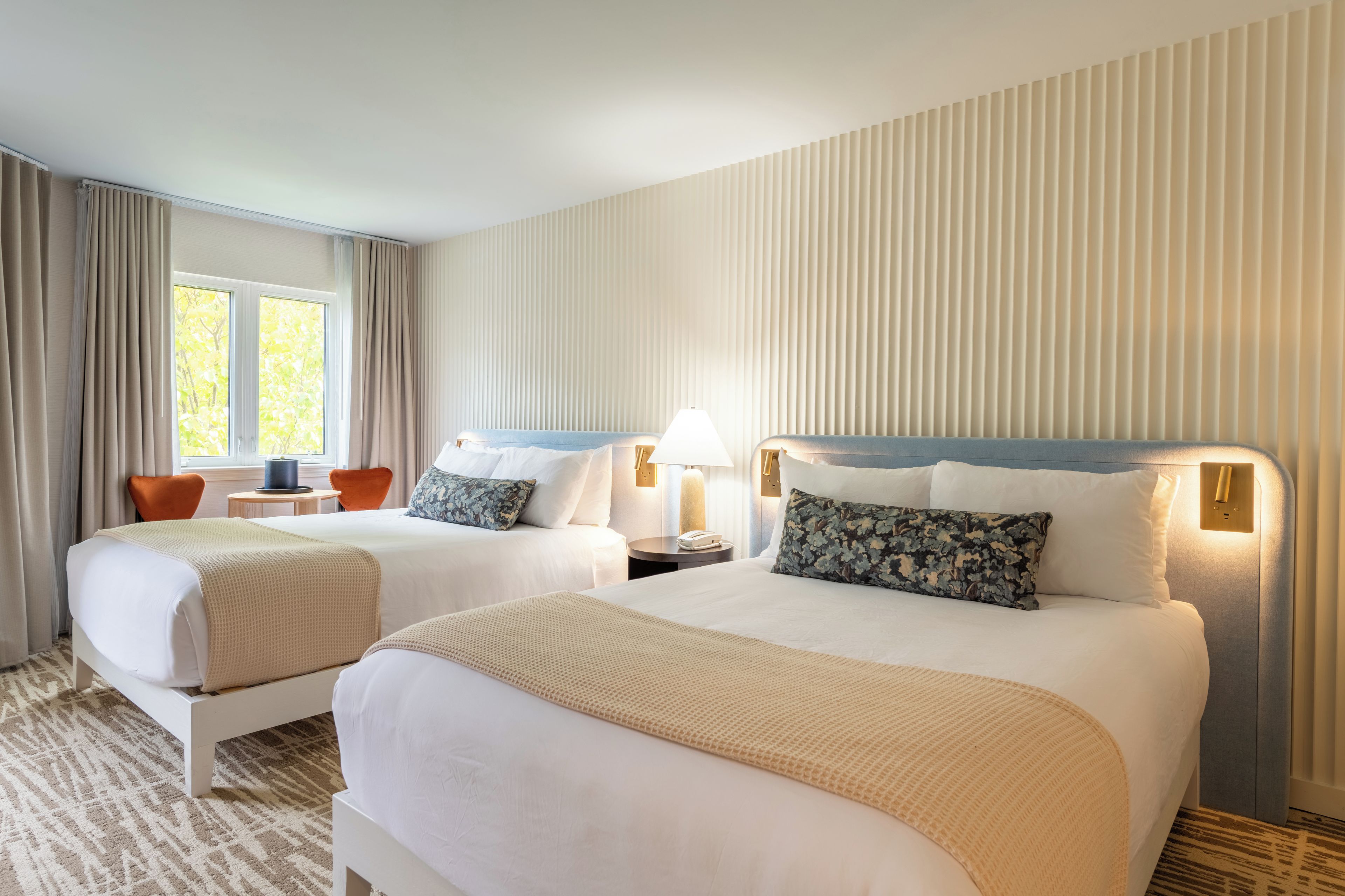 Quarto de hotel com duas camas, um espaço de estar com uma poltrona, luminária e janela com cortinas.