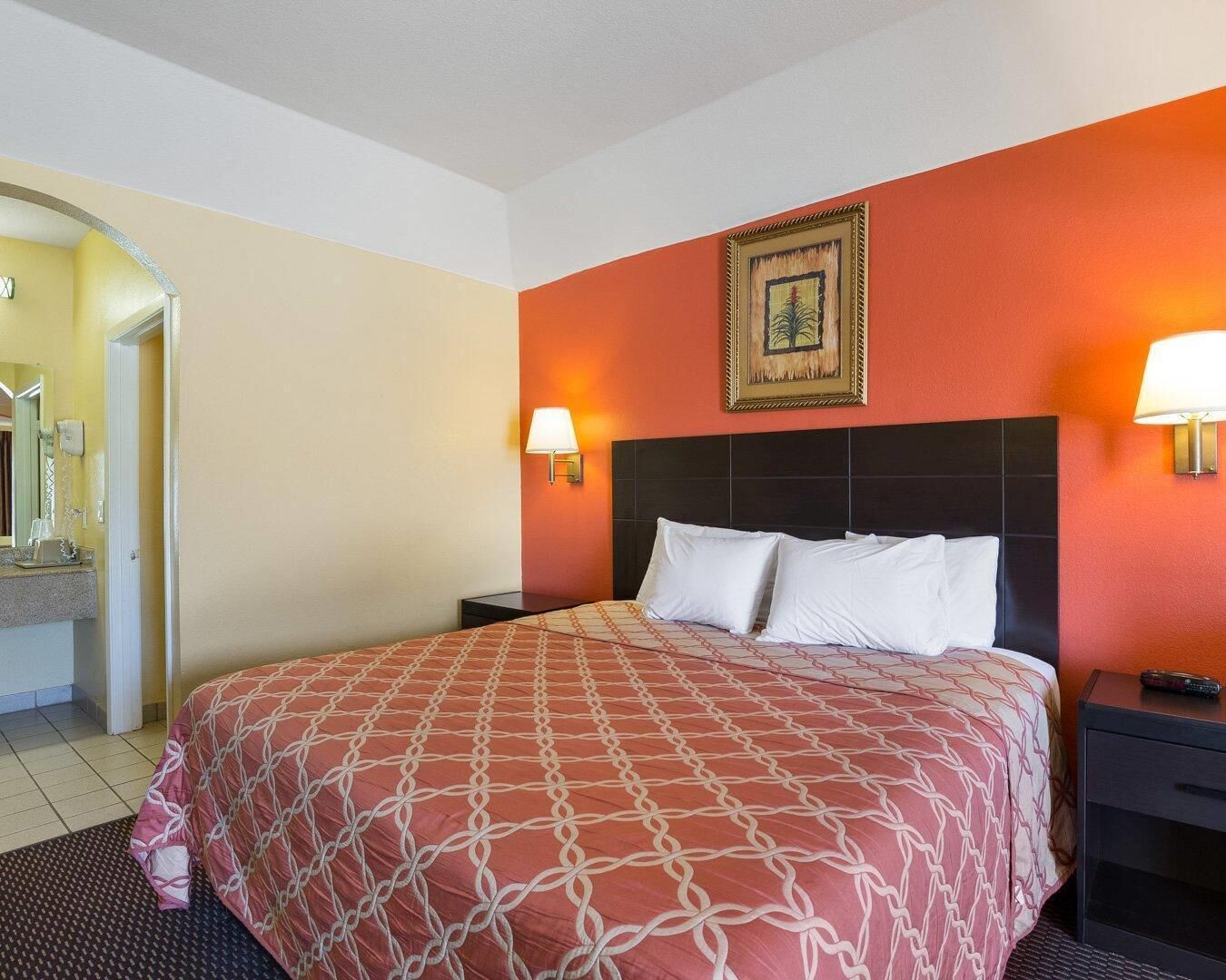 Quarto de hotel com uma cama grande, duas mesas de cabeceira, um criado-mudo e uma luminária fixada na parede, além de um quadro emoldurado sobre a cama.