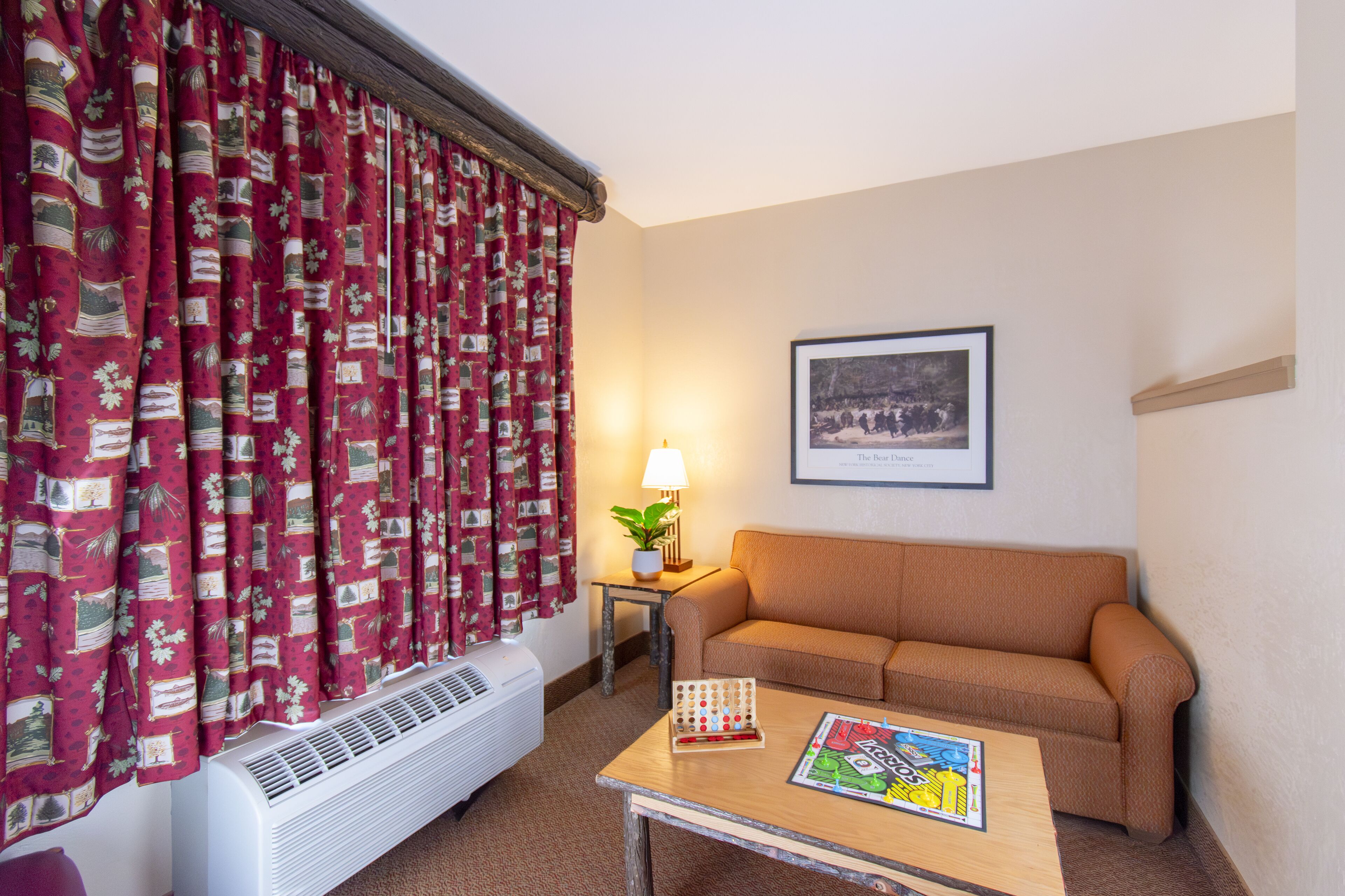 Um quarto aconchegante com uma cortina vermelha estampada, um sofá marrom, uma mesinha com um abajur, um quadro na parede e um ar condicionado branco.
