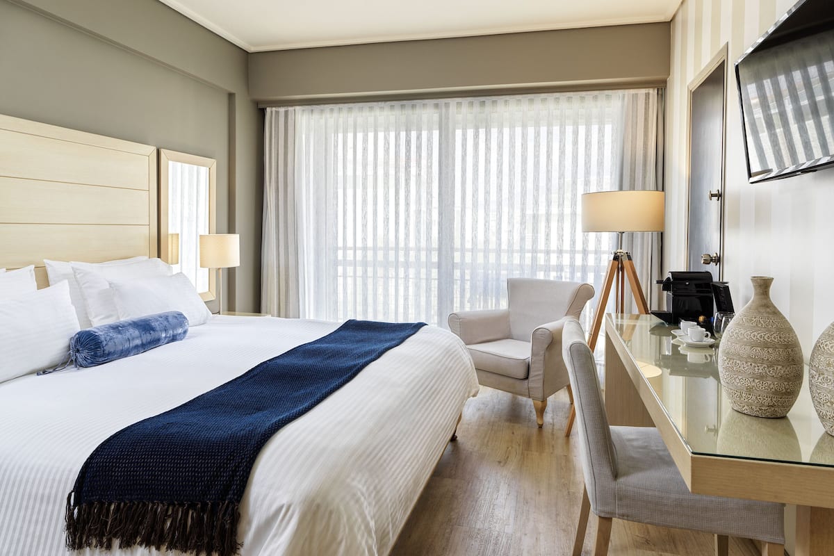 Quarto de hotel com cama, escrivaninha com luminária, cadeira e janela ampla com cortinas translúcidas.