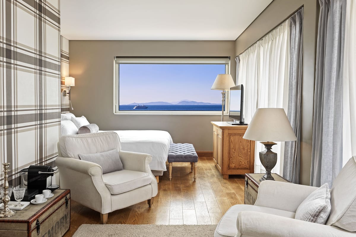 Quarto de hotel com uma janela grande, uma cama, duas poltronas, uma escrivaninha com uma luminária e uma mesinha com cafeteira.