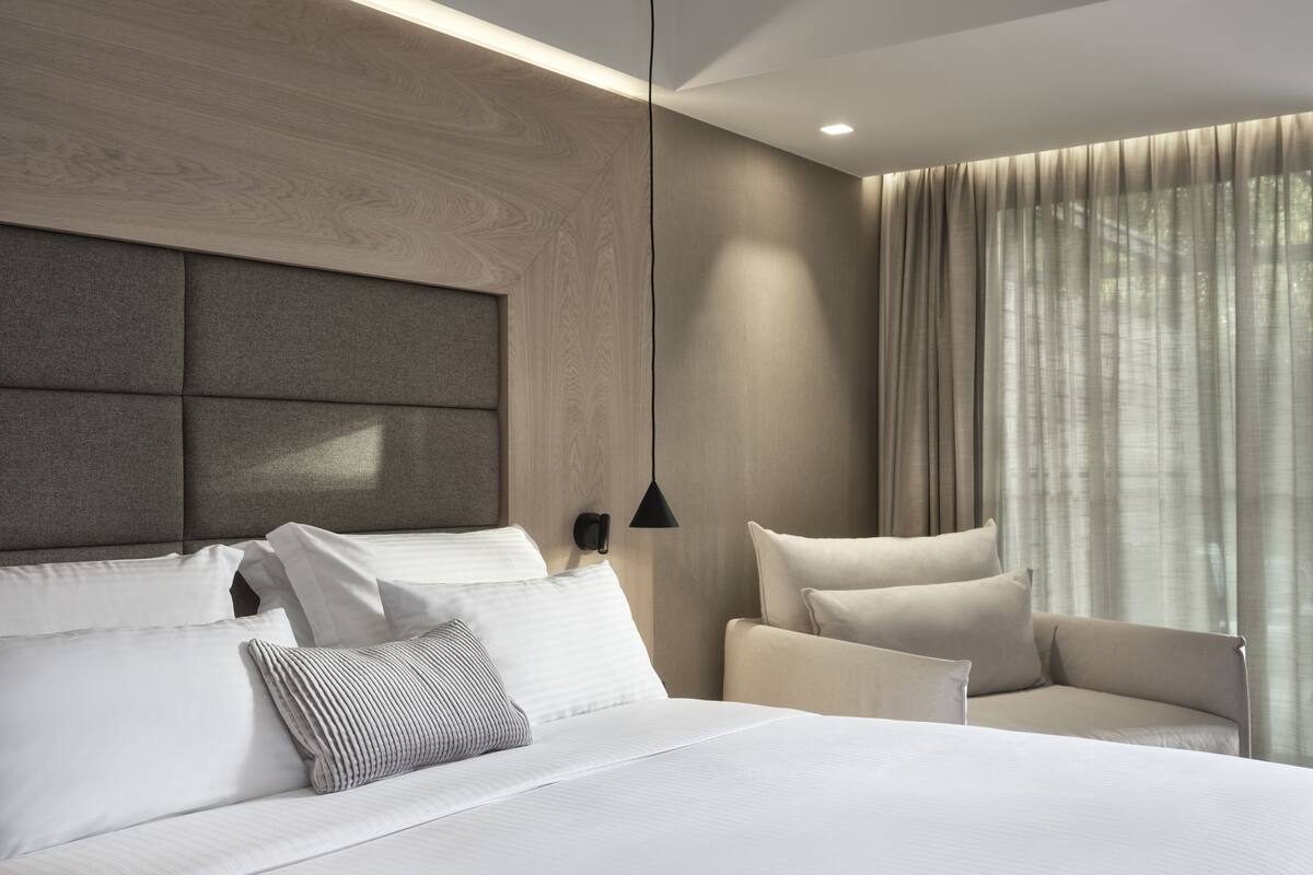 Quarto de hotel moderno com uma cama grande, cabeceira com design em painel de madeira, mesa de cabeceira com abajur e vista para o exterior através de cortinas finas.