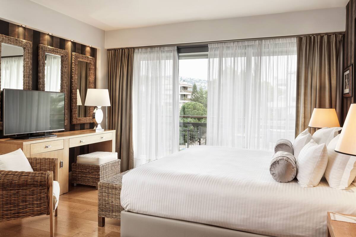 Quarto de hotel com uma cama grande, televisão, escrivaninha com abajur, mobiliário de vime e vista para a cidade.
