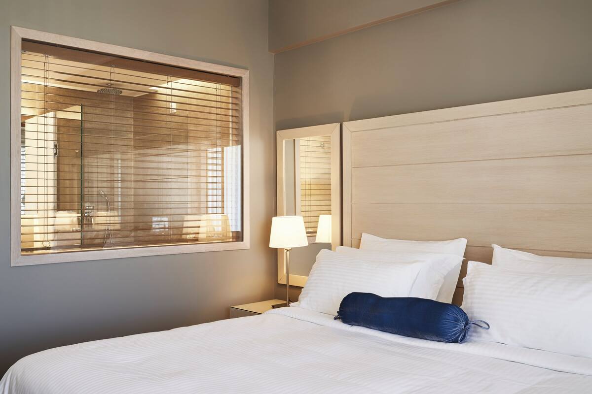 Quarto de hotel com uma grande janela, uma cama com roupa de cama branca, um abajur e uma cabeceira de madeira.