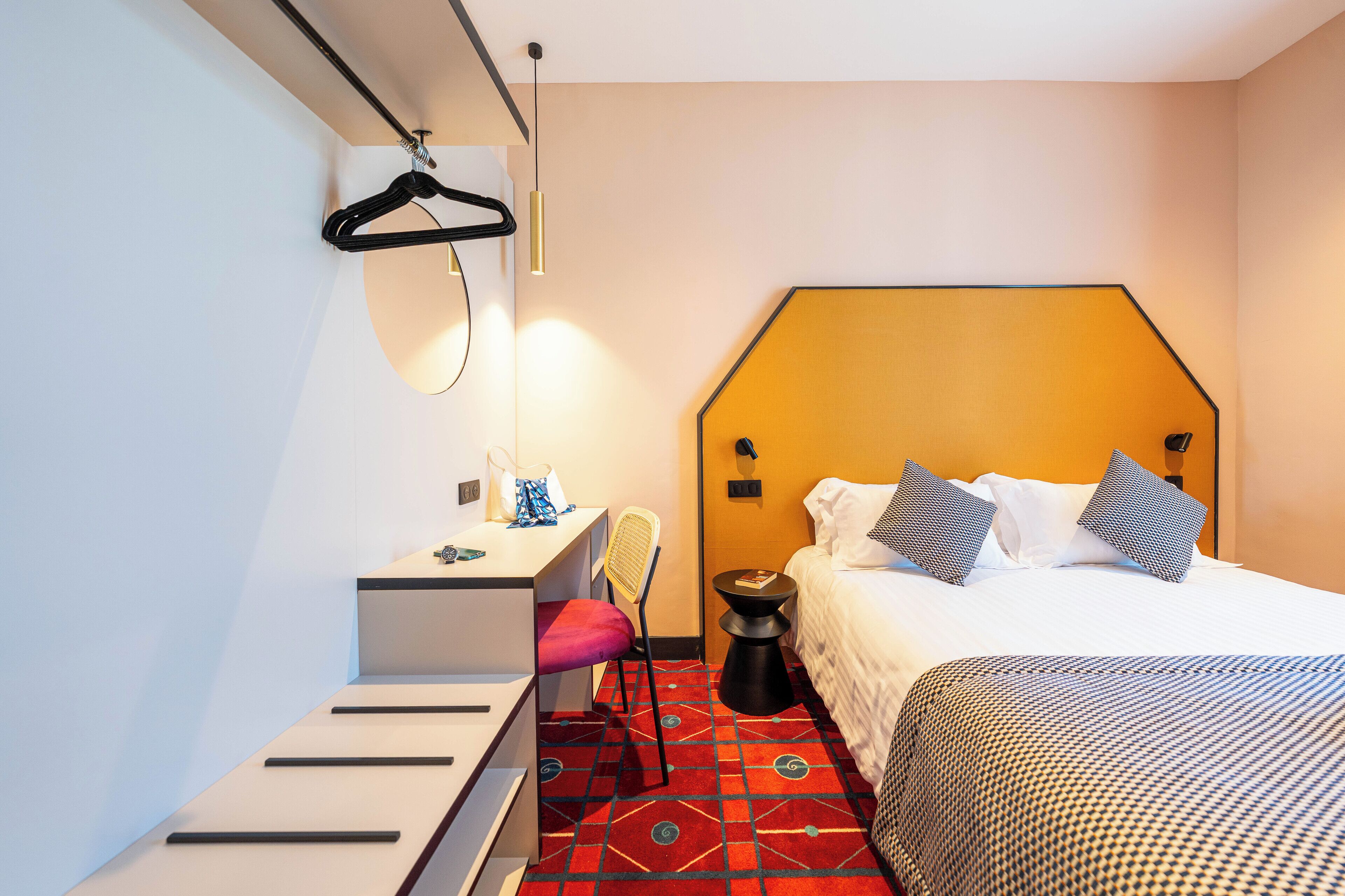 Quarto de hotel com cama, escrivaninha, cadeira e um cabideiro.