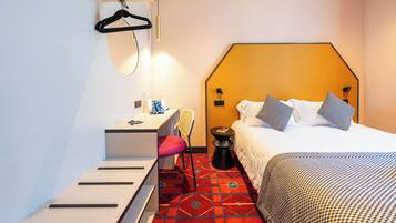 Quarto de hotel com cama, escrivaninha, cadeira e um cabideiro.