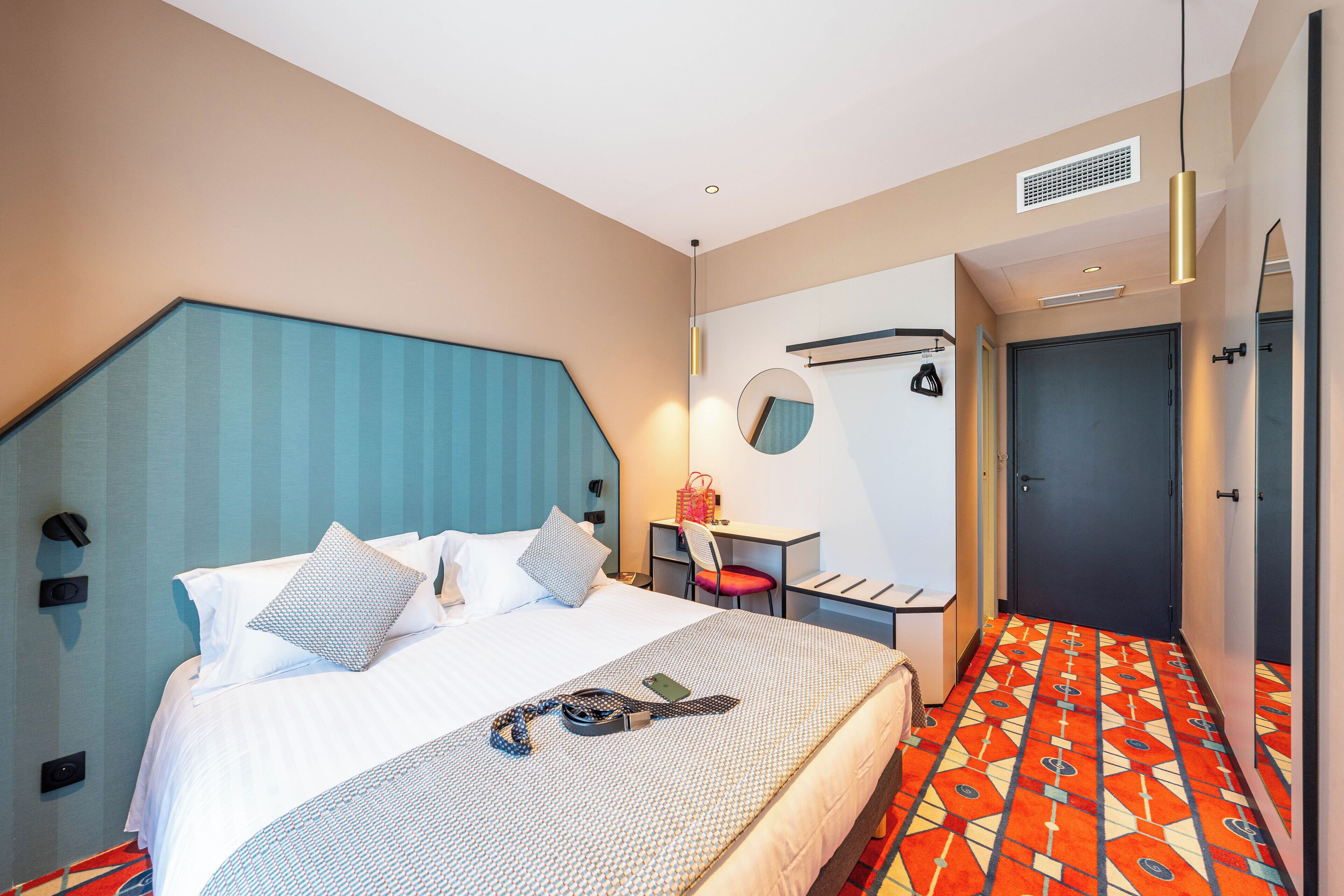Quarto de hotel com cama, escrivaninha, cadeira, espelho e porta.