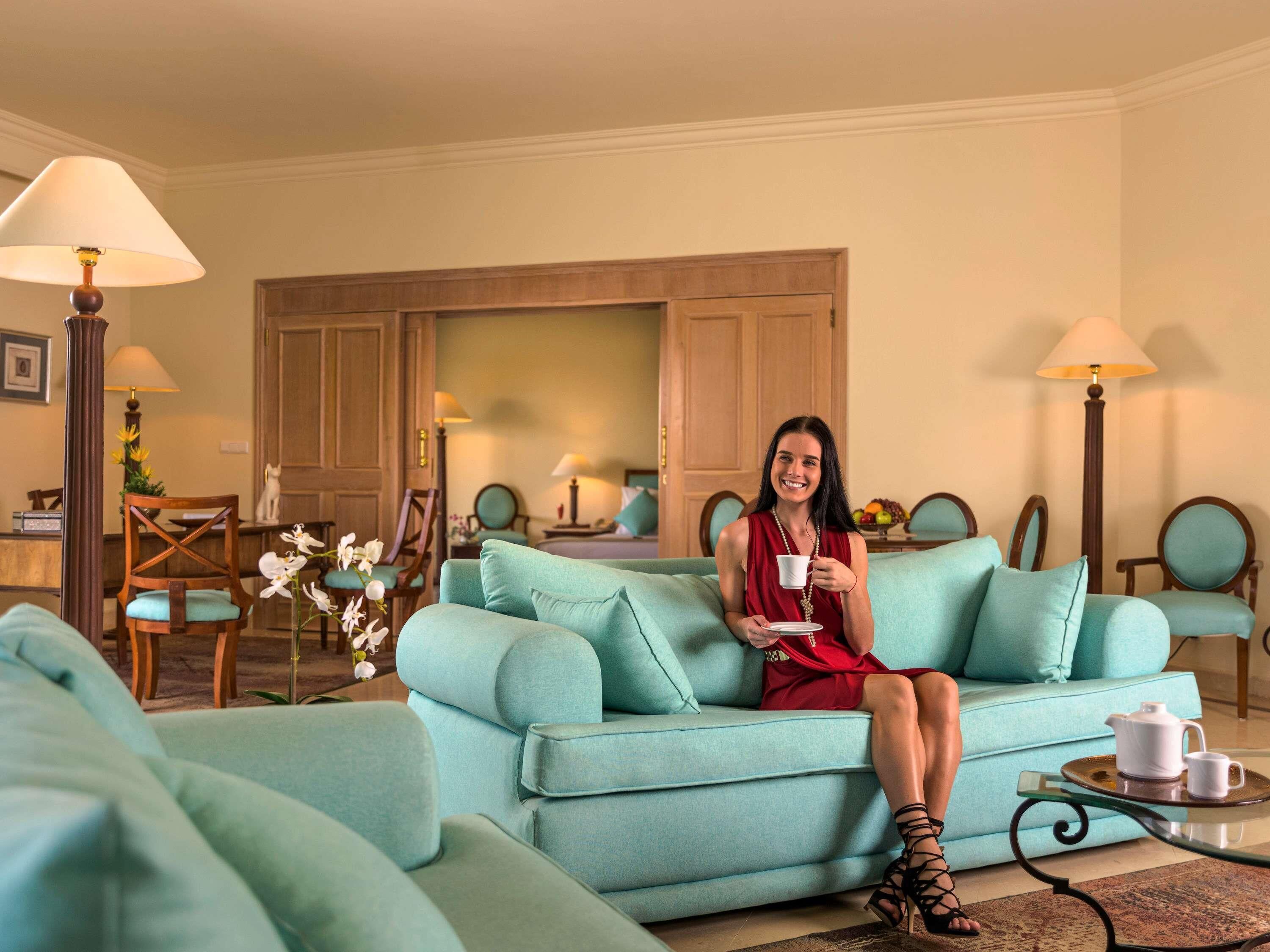 Uma mulher sentada em um sofá azul-turquesa em um quarto bem iluminado, com móveis de madeira, uma área de jantar ao fundo e uma mesa com um bule e uma xícara.