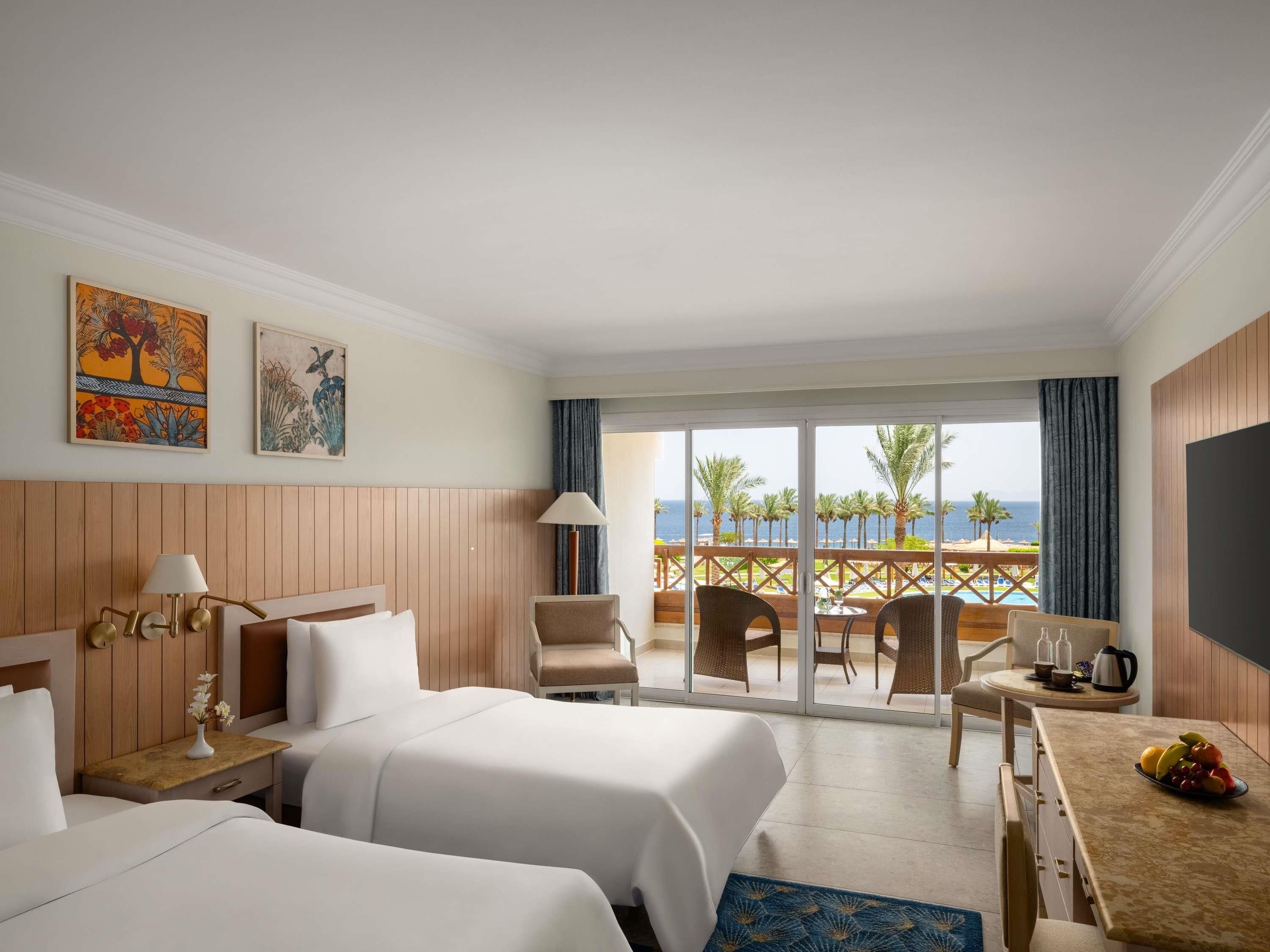 Quarto de hotel com duas camas, uma escrivaninha e varanda com vista para o mar.