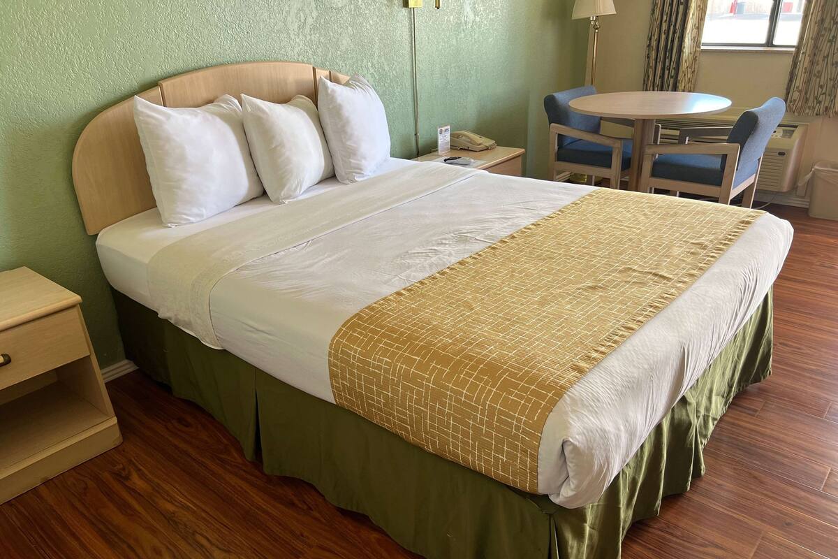 Quarto de hotel com uma cama grande, uma mesa de cabeceira, escrivaninha com cadeira, luminária e janela com cortinas.