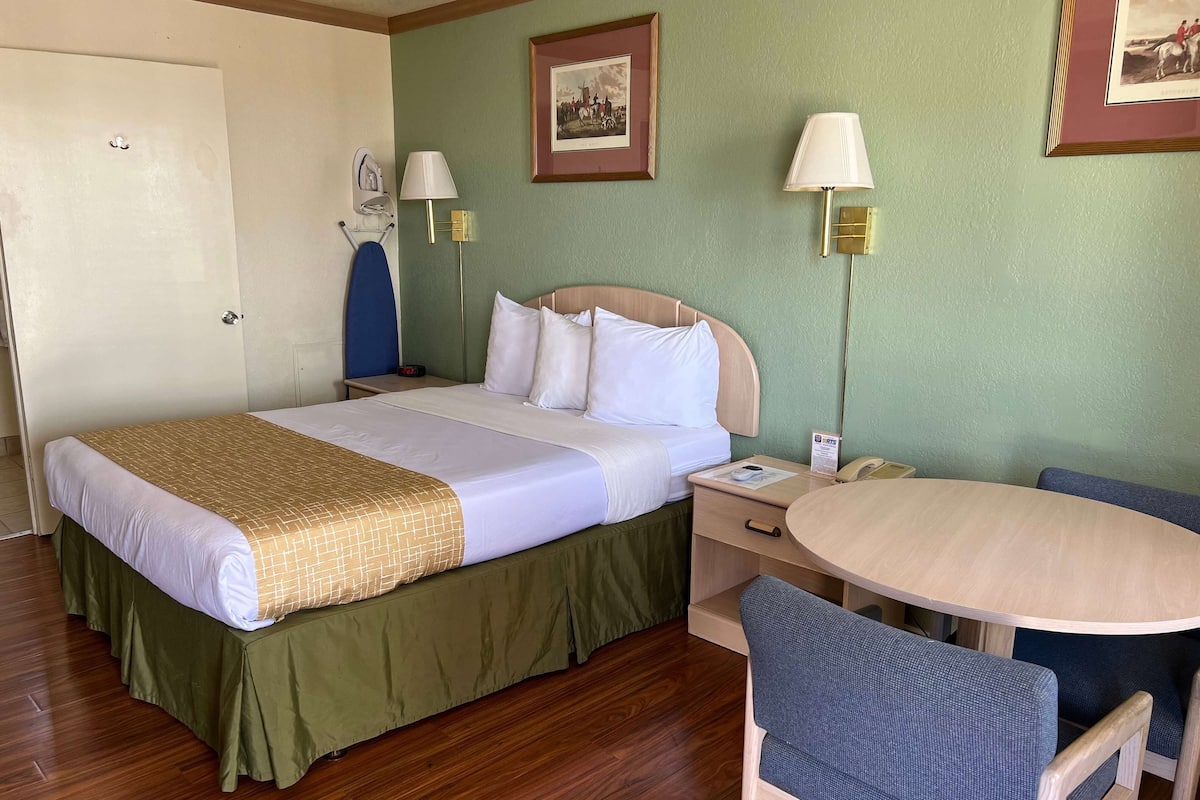 Quarto de hotel com uma cama, uma escrivaninha com candeeiro, uma cadeira e uma mesinha.
