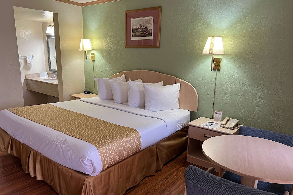 Quarto de hotel com cama, mesa de cabeceira, abajur e uma pequena mesa redonda.