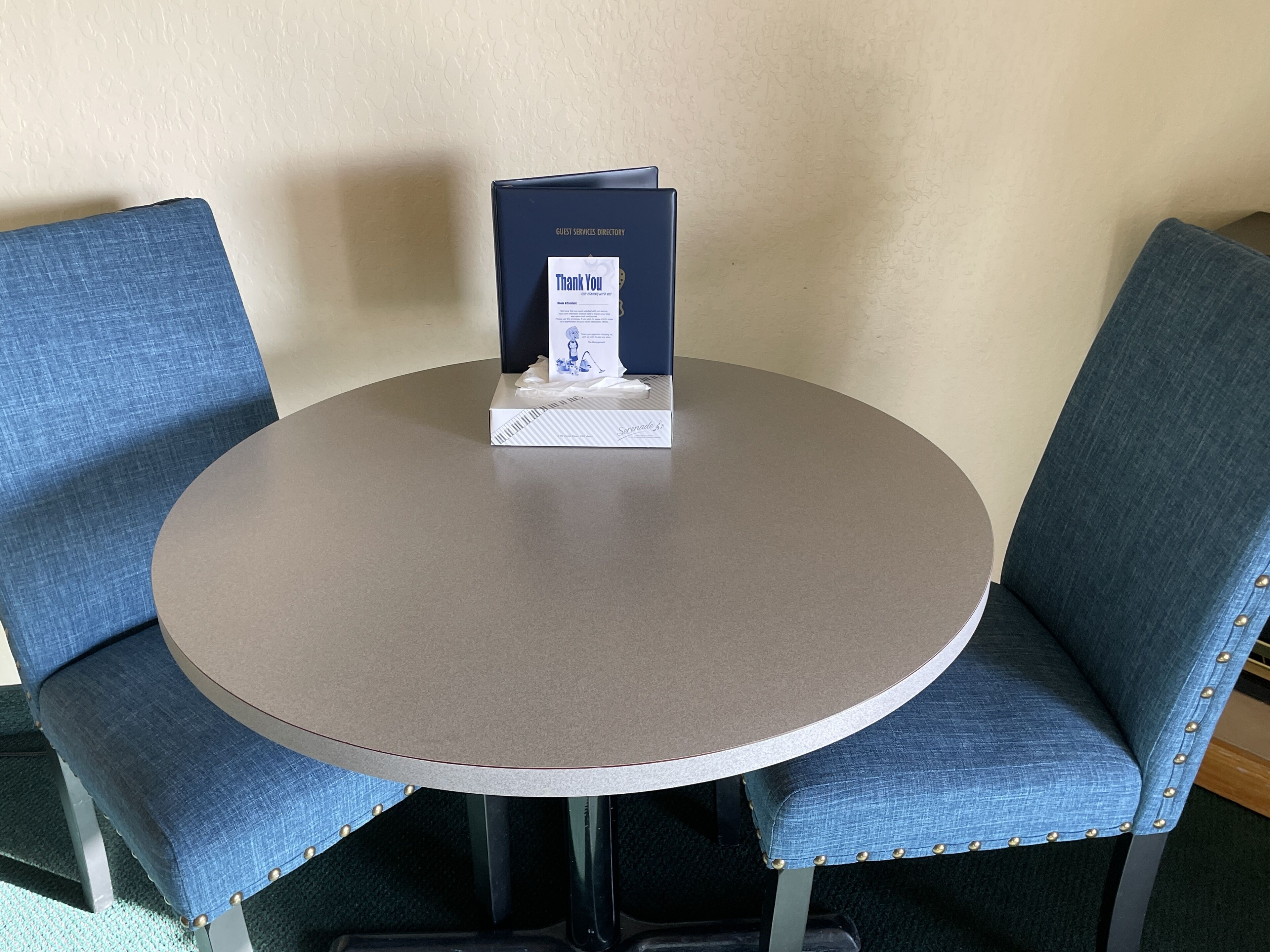 Uma mesa redonda com um guia azul e uma caixa de lenços, acompanhada por duas cadeiras estofadas azuis com detalhes em metal.