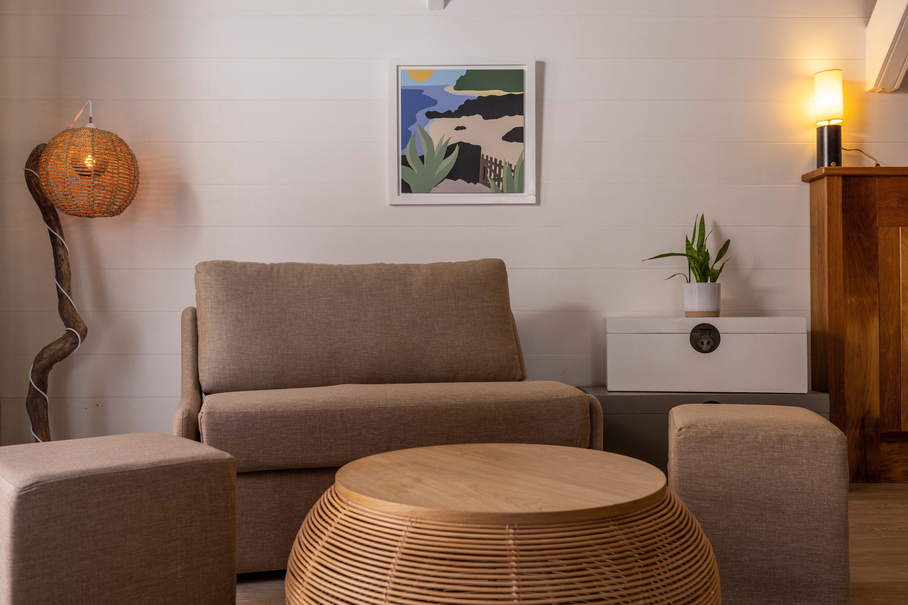 Uma sala de estar moderna com sofá, dois pufes, uma mesa de centro redonda de vime, um abajur fixado na parede, um quadro emoldurado, uma pequena planta em vaso e um armário de madeira.