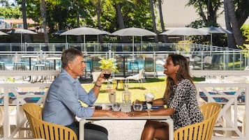 Um casal jantando em uma mesa ao ar livre com vista para a piscina, cercado por guarda-sóis e cadeiras.