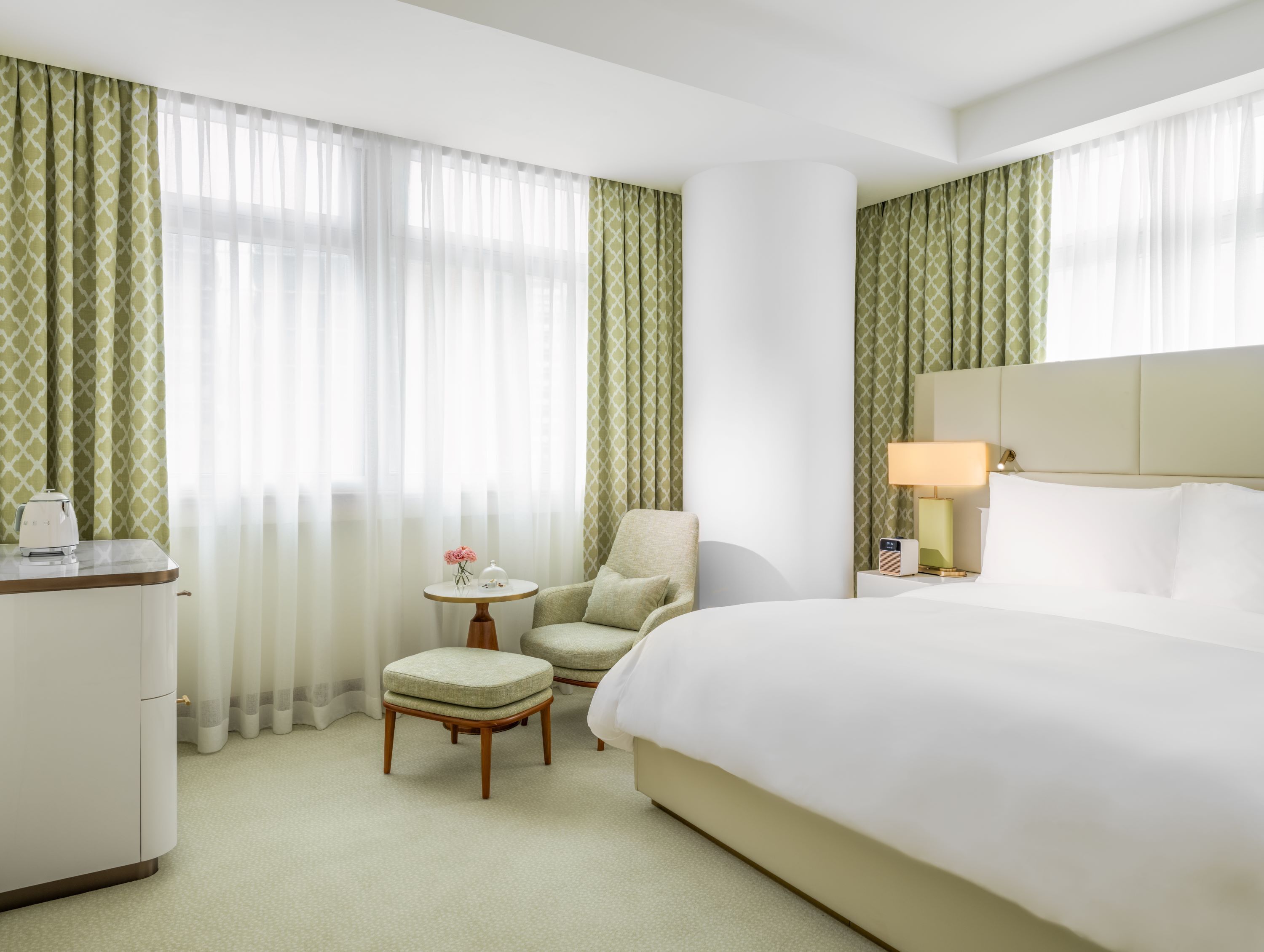 Quarto de hotel com cama, uma cadeira, uma pequena mesa com abajur e escrivaninha com chaleira.