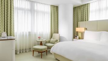 Quarto de hotel com cama, uma cadeira, uma pequena mesa com abajur e escrivaninha com chaleira.