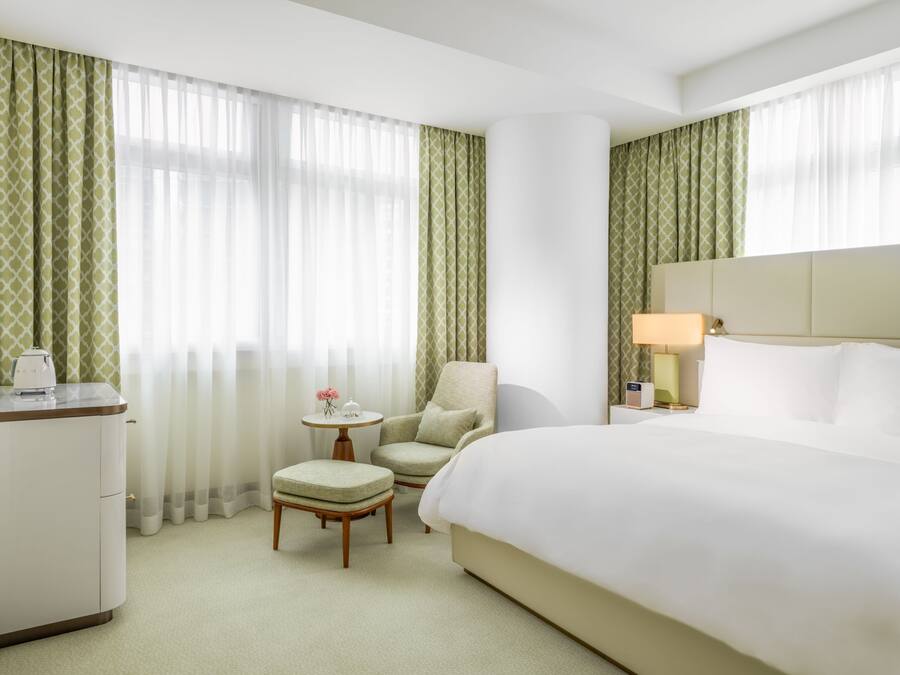 Quarto de hotel com cama, uma cadeira, uma pequena mesa com abajur e escrivaninha com chaleira.